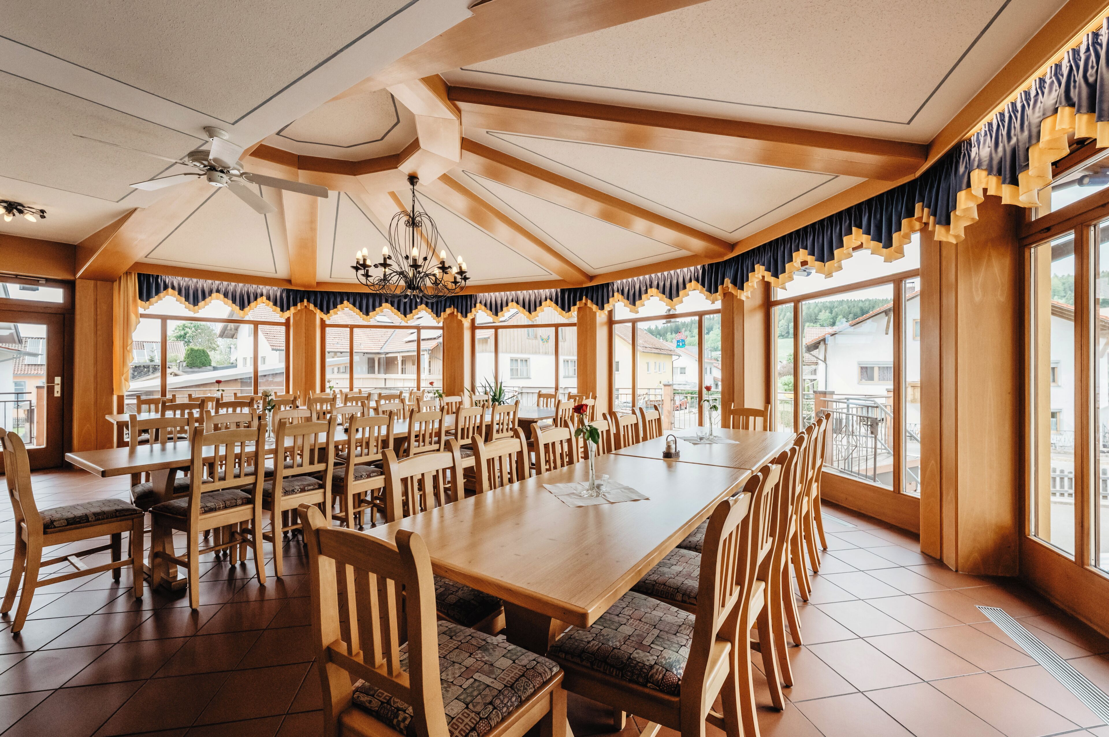 Uma sala de jantar espaçosa com mesas e cadeiras de madeira, janelas amplas e um ventilador de teto.