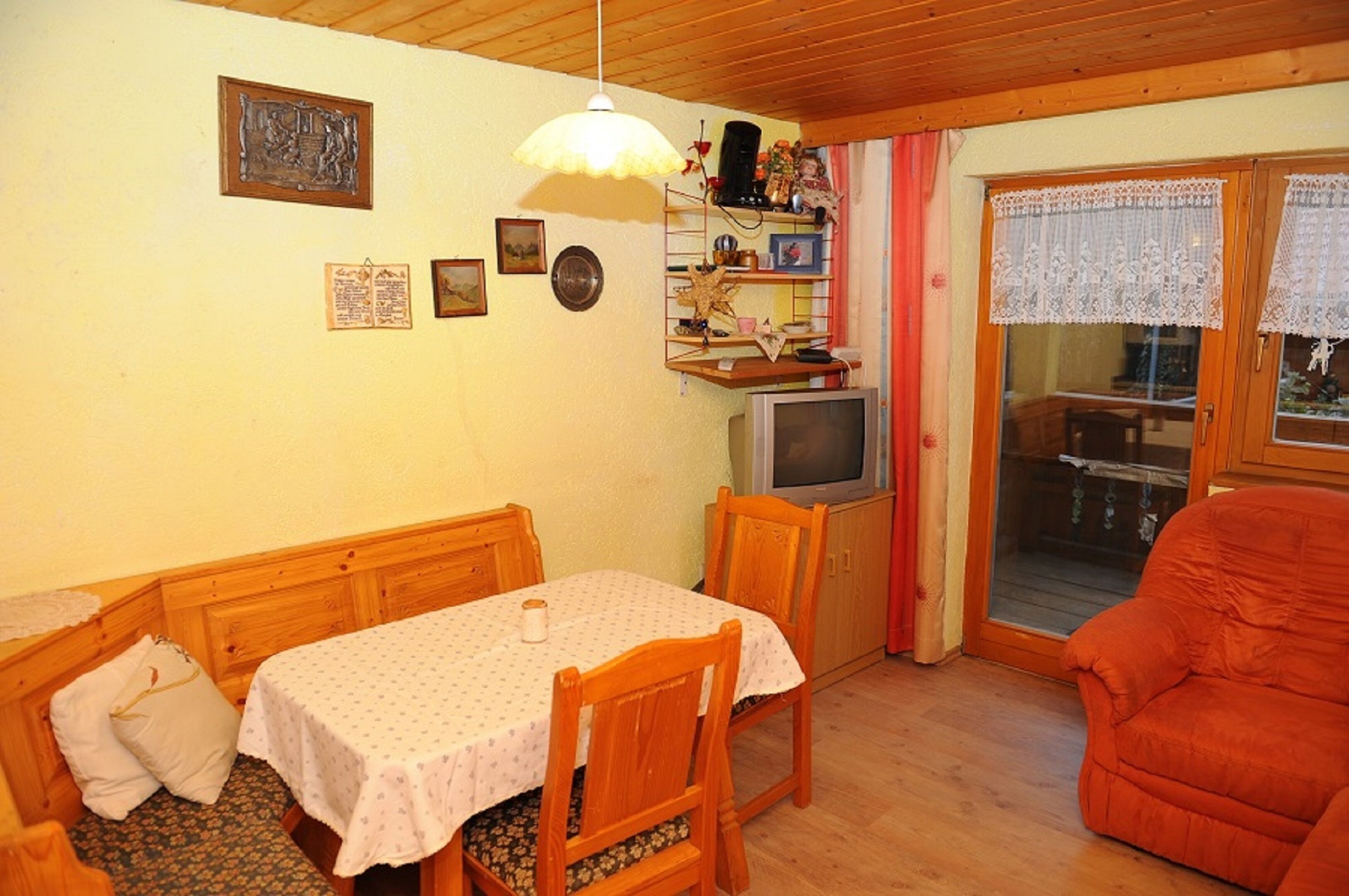 Quarto aconchegante com um banco de madeira, uma mesa com toalha branca, uma televisão e uma poltrona vermelha.