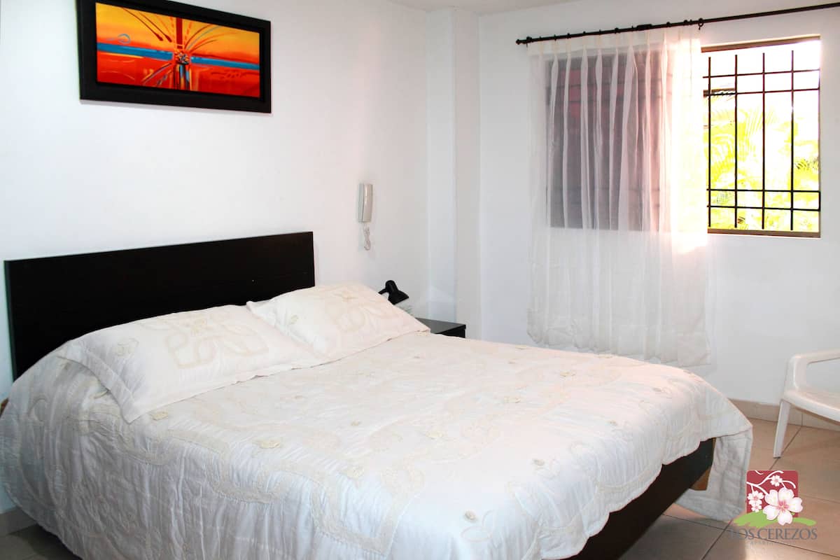 Uma cama bem arrumada com colcha branca e travesseiros, cabeceira preta, mesa de cabeceira com abajur, janela com cortina e um quadro na parede.