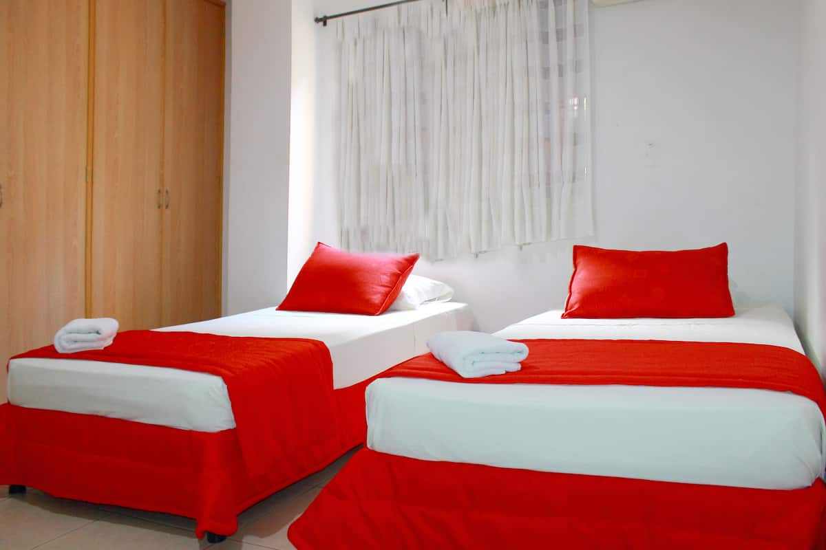 Duas camas de solteiro com roupa de cama vermelha e almofadas brancas, um guarda-roupa de madeira e uma janela com cortinas brancas.