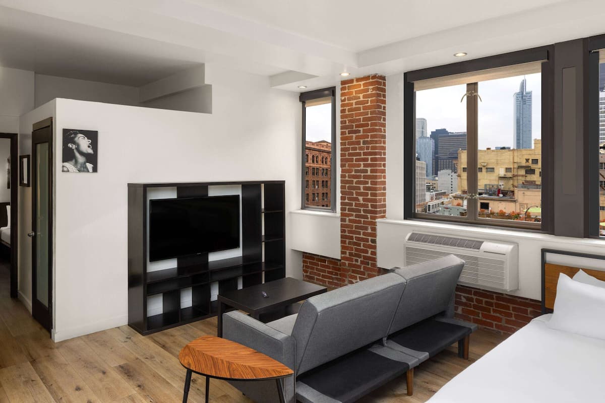 Quarto de hotel moderno com televisão grande, sofá cinzento, mesa de madeira pequena e vista para a cidade através de amplas janelas.