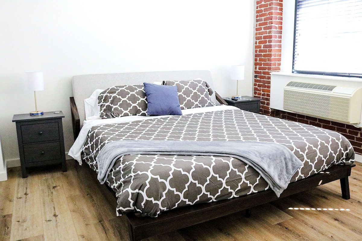 Quarto com colcha estampada, mesa de cabeceira com abajur e janela com parede de tijolos.