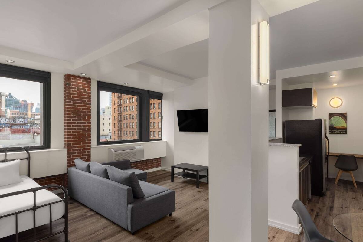 Apartamento moderno com sala de estar que inclui um sofá cinzento, uma poltrona branca e uma pequena mesa. Há uma cozinha com frigorífico, pia e mesa de jantar. Grandes janelas oferecem vista para a paisagem urbana.