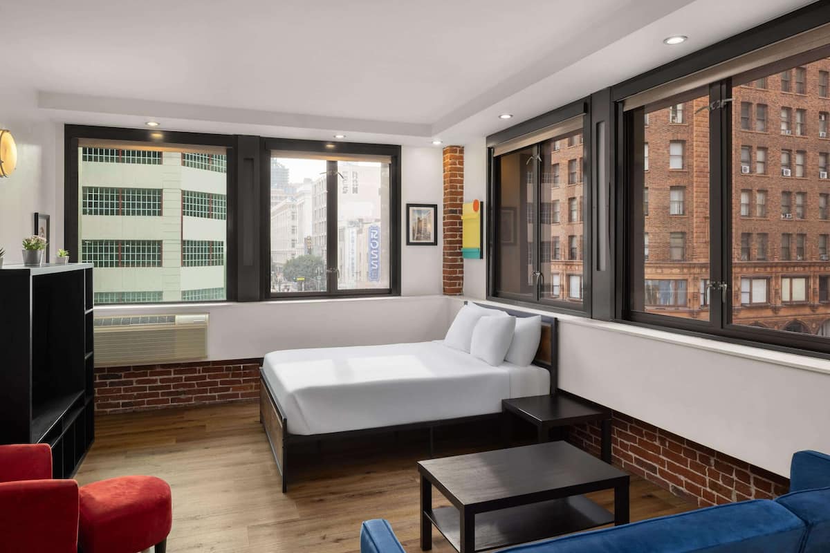 Quarto de hotel moderno com uma cama grande, área de estar com uma cadeira vermelha e um sofá azul, uma mesinha e amplas janelas com vista para a cidade.