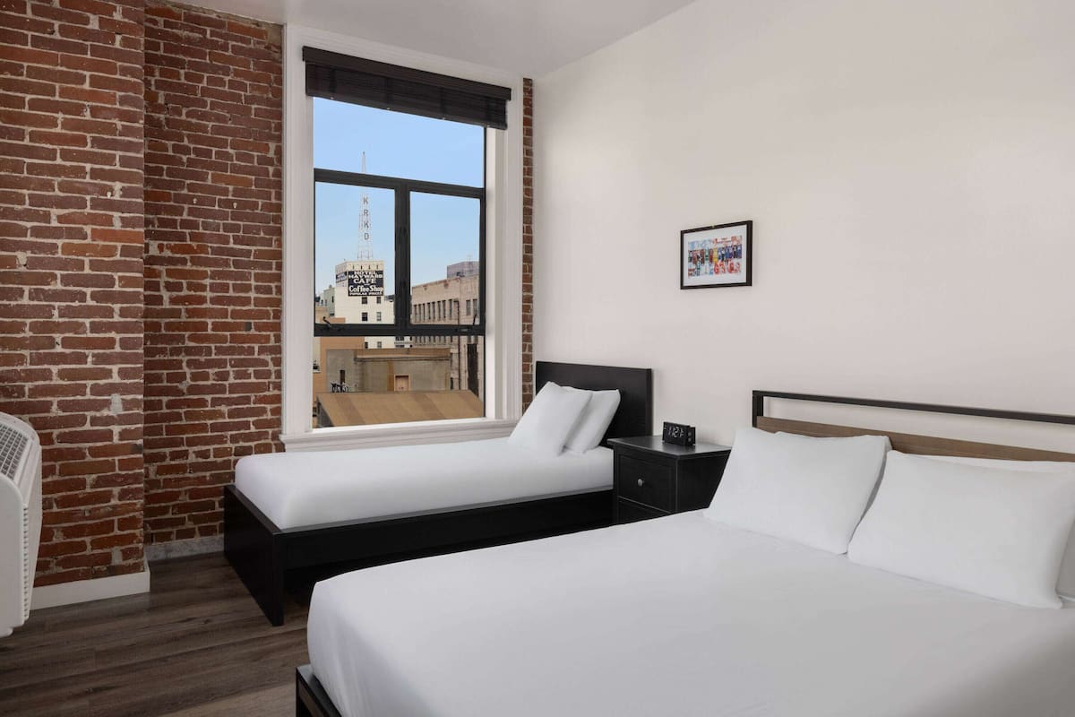 Quarto de hotel com parede de tijolos, duas camas, janela com vista para edifícios, uma mesa pequena com um relógio e um quadro pendurado na parede.