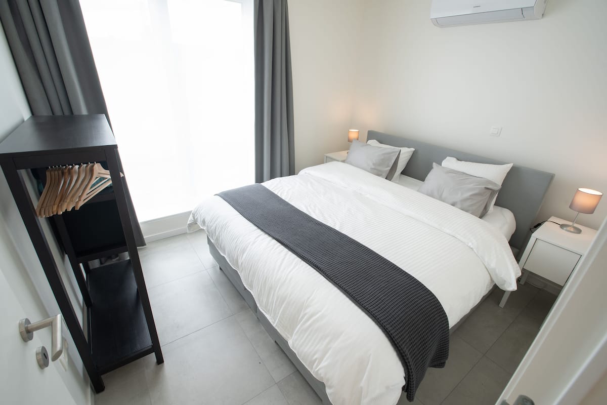 Quarto de hotel bem arrumado, com cama, mesas de cabeceira, guarda-roupa e janela com cortinas.