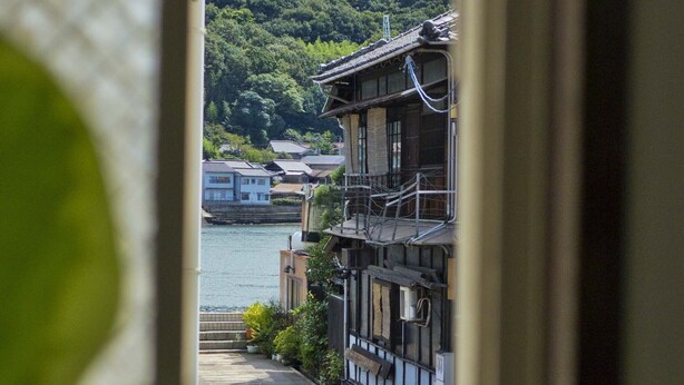 Visão pela janela de um edifício japonês tradicional, com fachada de madeira, um pequeno jardim e um corpo d'água ao fundo.