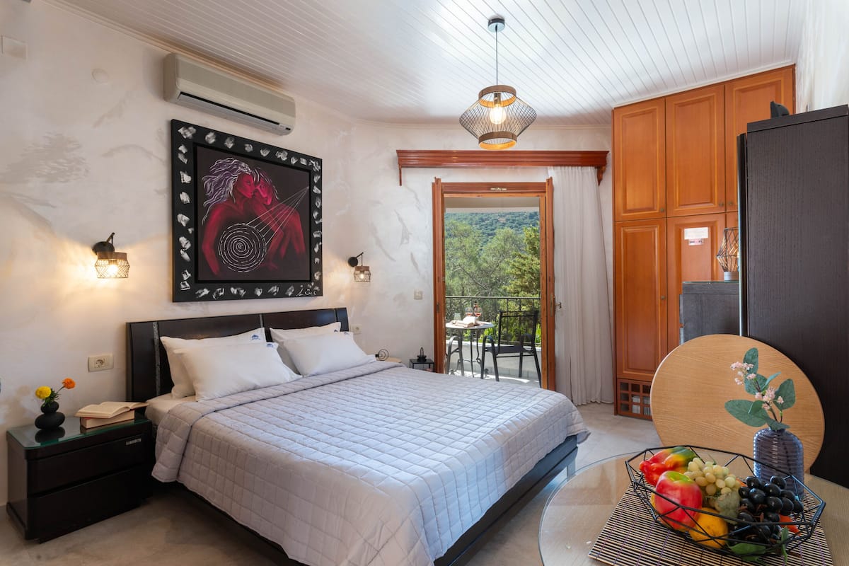 Quarto com cama, mesas de cabeceira, ar condicionado fixado na parede, uma obra de arte emoldurada, luminária pendente e vista para o exterior por uma porta.