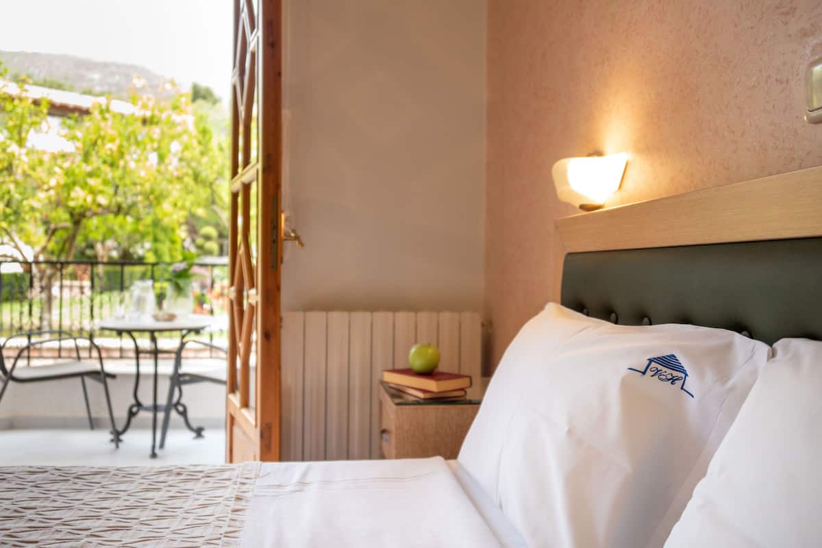 Quarto de hotel com uma cama, mesa de cabeceira com um livro e uma maçã, abajur e vista para área externa com mesa e cadeiras.
