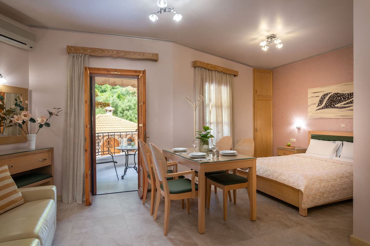 Quarto de hotel com cama, mesa de jantar com cadeiras, mesinha com um vaso de flores e varanda com vista para o verde.
