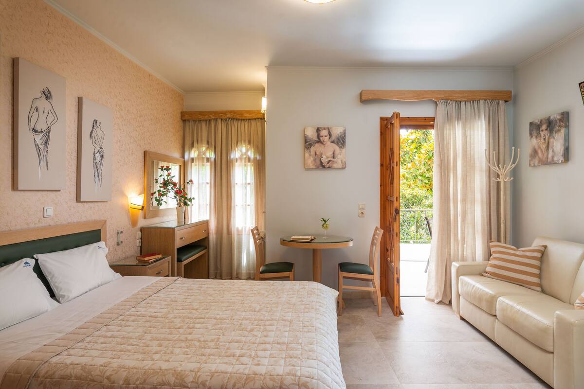 Quarto de hotel com cama, sofá, mesa de jantar com cadeiras, mesinha com um vaso e janela com cortinas.