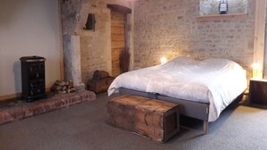 Quarto rústico com parede de pedra, cama de madeira, um banquinho e uma pequena prateleira com um abajur.
