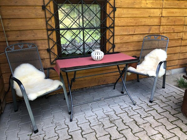 Uma cabana de madeira com uma pequena área de varanda, com duas cadeiras de metal com almofadas de pele, uma mesa de metal e um vaso decorativo branco.