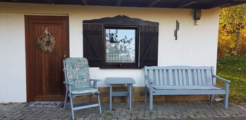 Uma porta de madeira com uma guirlanda, uma cadeira estampada, uma mesinha e um banco azul em frente a um edifício com uma janela e um candeeiro fixado na parede.