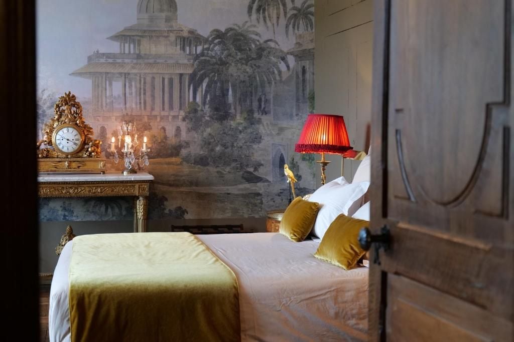 Quarto com cama, mesa de cabeceira, relógio, luminária e um mural na parede.