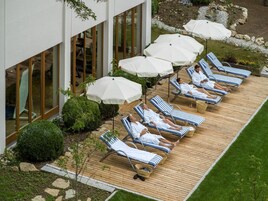 Um deck com espreguiçadeiras e guarda-sóis, cercado por um jardim e um edifício com grandes janelas.