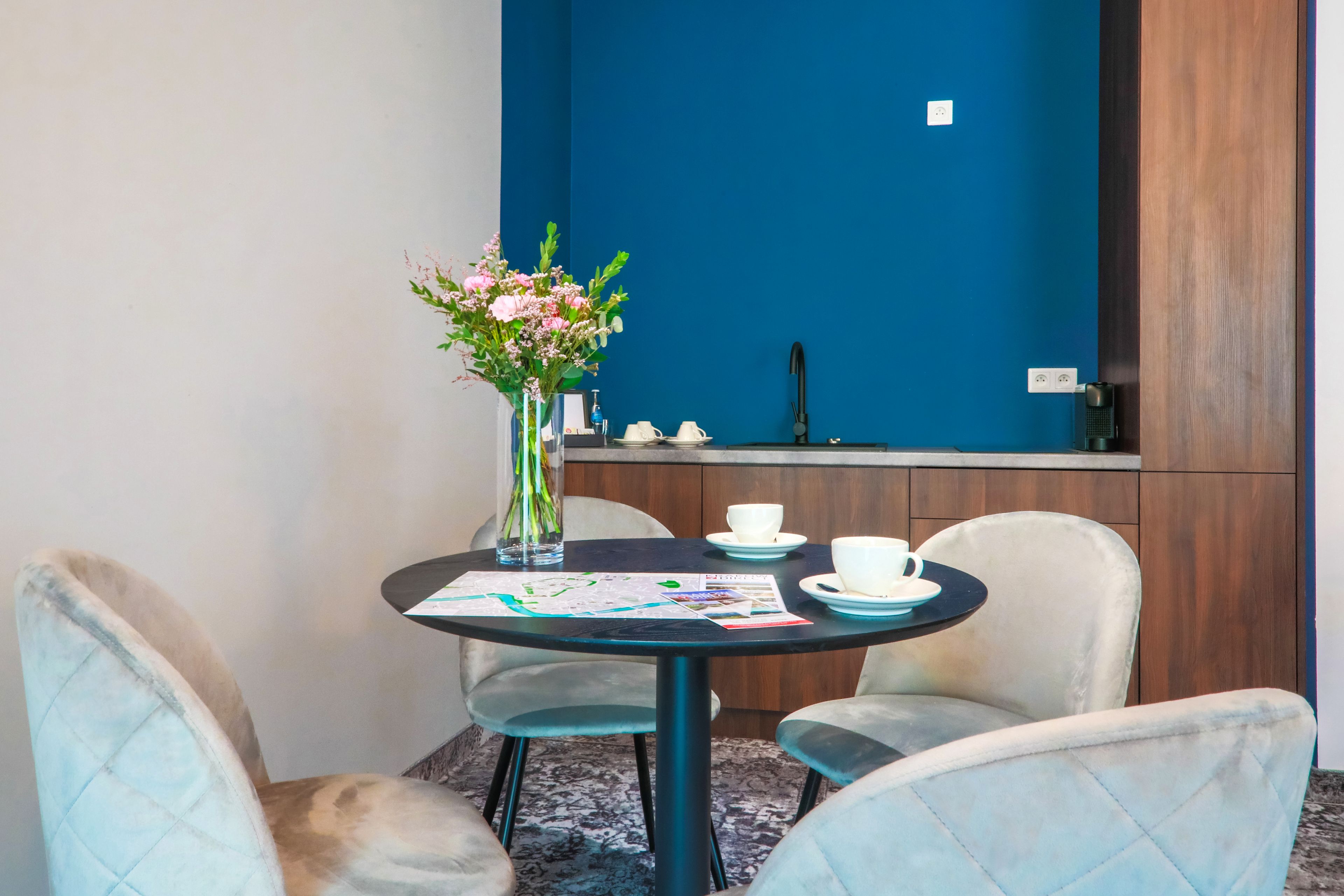 Quarto de hotel com mesa redonda, duas cadeiras, um vaso com flores, um cantinho para café e uma parede azul.
