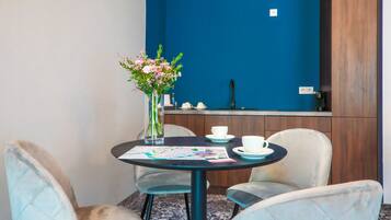 Quarto de hotel com mesa redonda, duas cadeiras, um vaso com flores, um cantinho para café e uma parede azul.