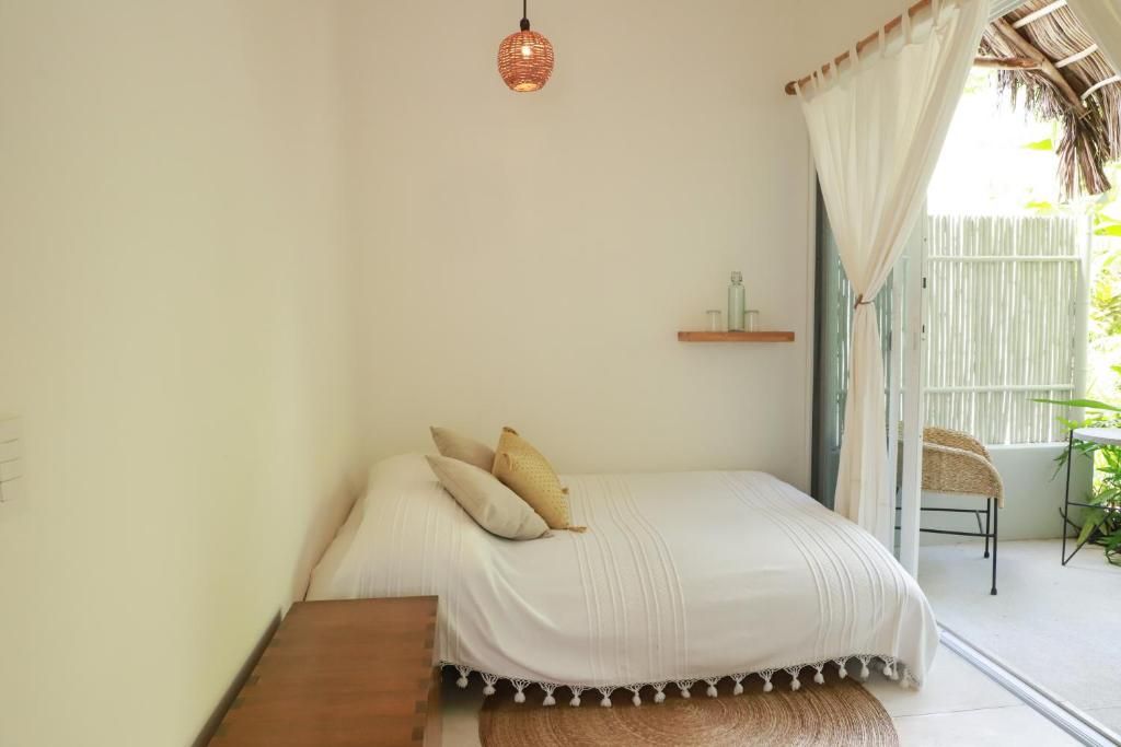 Quarto com cama, mesa de cabeceira, luminária pendente, janela com cortinas e uma pequena prateleira com garrafas.