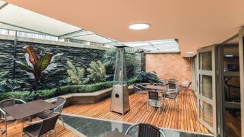 Área de varanda externa com piso de madeira, cadeiras de metal, mesa de vidro e parede de pedra com plantas.