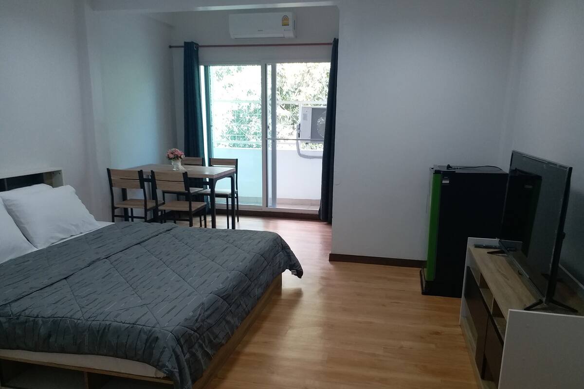Quarto com cama, escrivaninha, cadeira, televisão e porta de vidro deslizante para uma varanda.