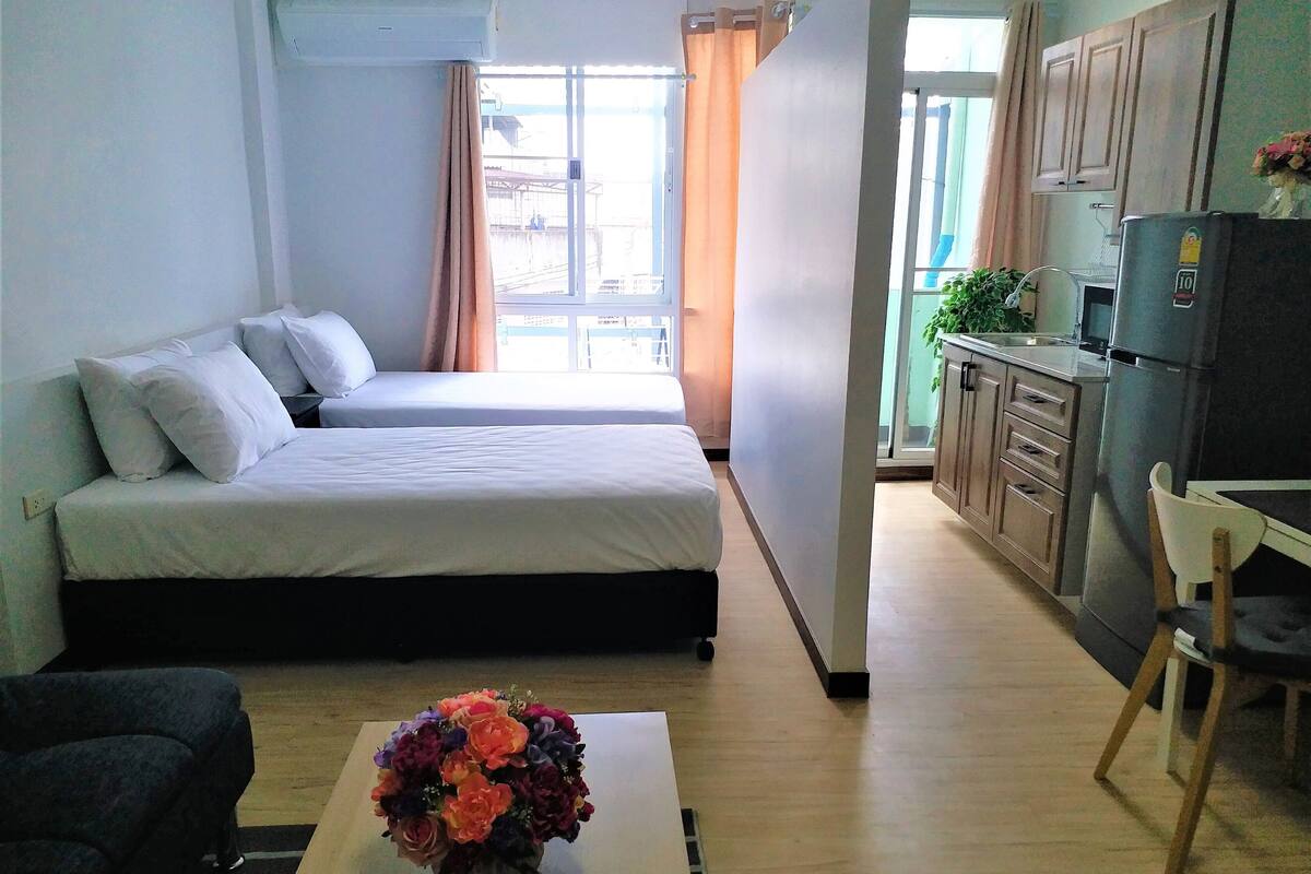 Quarto com duas camas, uma mesinha com um vaso de flores e uma kitchenette com frigorífico e armários.