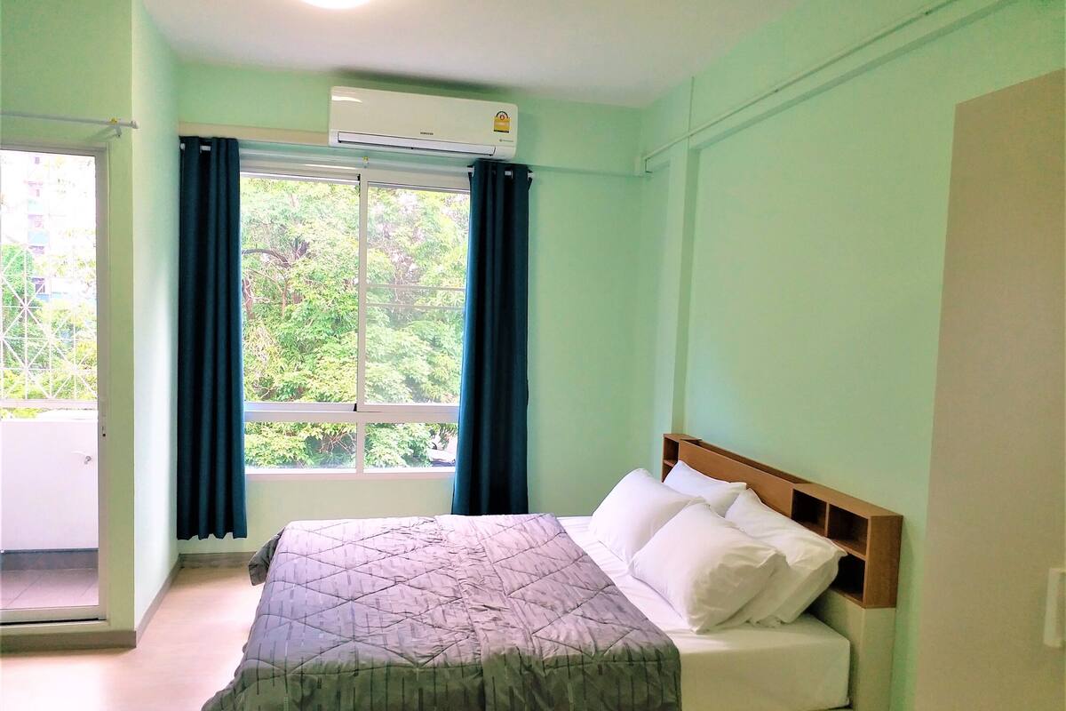Quarto com cama, janela com cortinas, ar condicionado e vista para o verde lá fora.