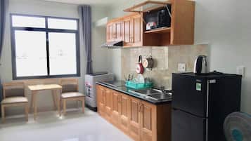 Cozinha compacta com armários de madeira, geladeira preta, micro-ondas e ventilador. Há uma janela com cortinas e uma mesa com cadeiras.