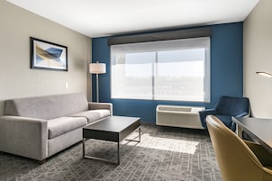 Quarto de hotel moderno com um sofá cinzento, uma mesa pequena, uma poltrona azul e uma grande janela com vista para o exterior.