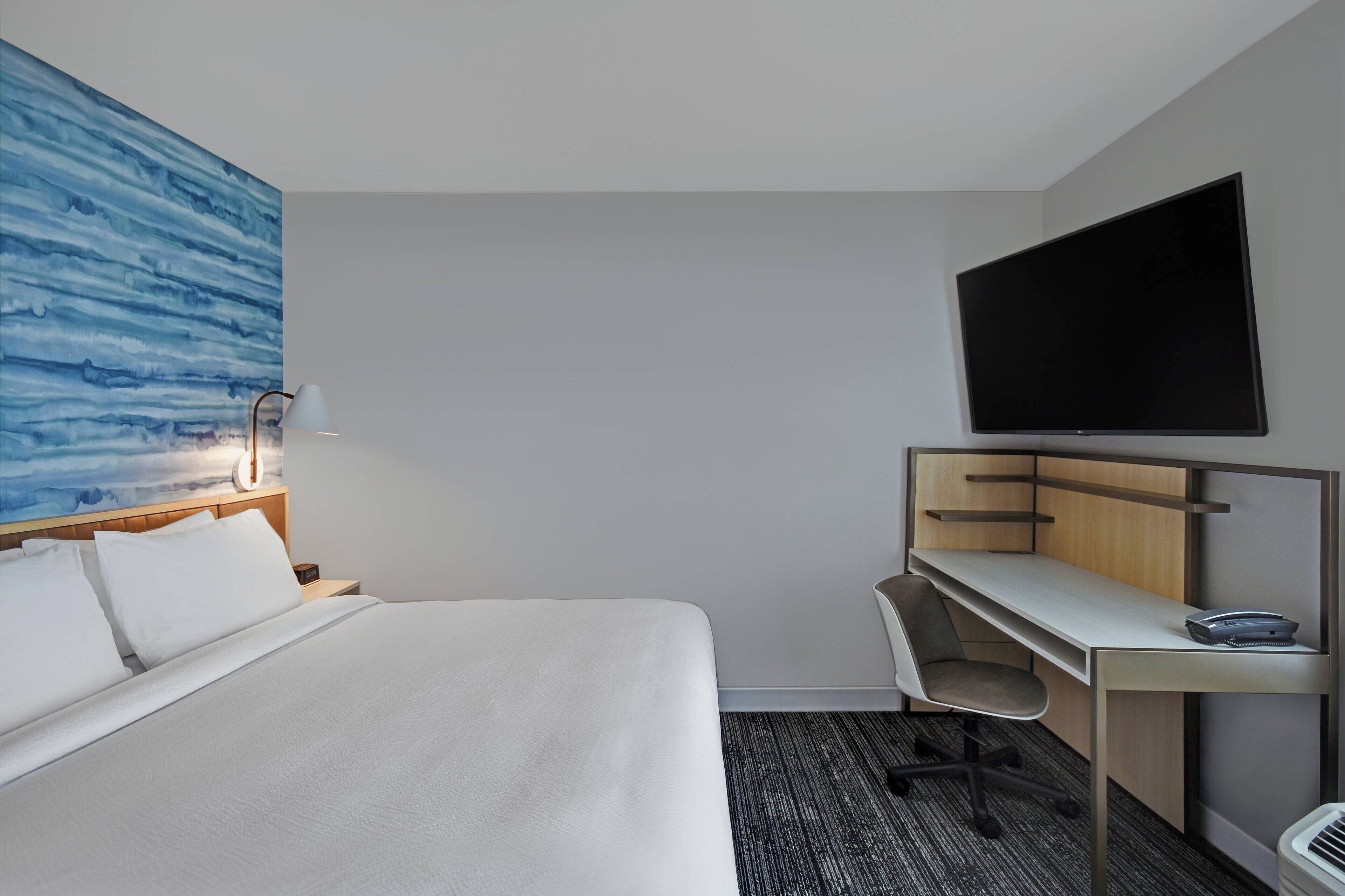 Quarto de hotel com uma cama, uma escrivaninha, uma cadeira, televisão de tela plana e um quadro na parede.