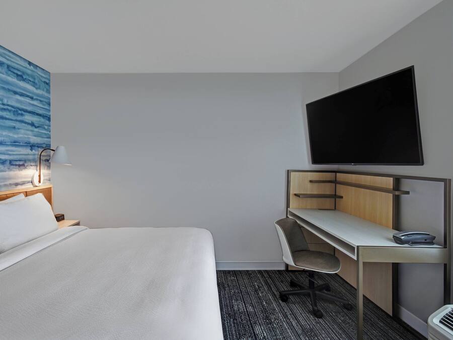 Quarto de hotel com uma cama, uma escrivaninha, uma cadeira, televisão de tela plana e um quadro na parede.
