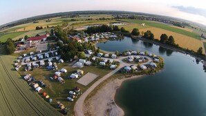 Vista aérea de um camping com veículos recreativos e tendas, um lago e campos ao redor.