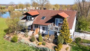Uma casa de dois andares com telhado de telhas vermelhas, varanda e vista para um lago.