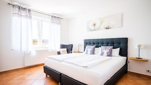 Cama bem arrumada com cabeceira escura, uma cadeira, mesa de cabeceira com abajur e janela com cortinas translúcidas.
