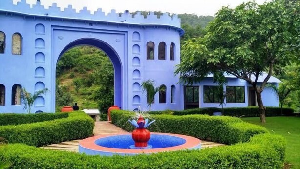Um edifício azul grande com entrada em arco, cercado por sebes bem cuidadas e uma fonte central.