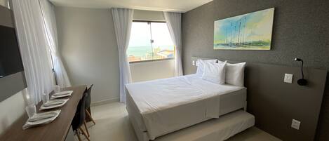 Quarto de hotel com cama, escrivaninha com cadeiras, um quadro na parede e janela com cortinas.