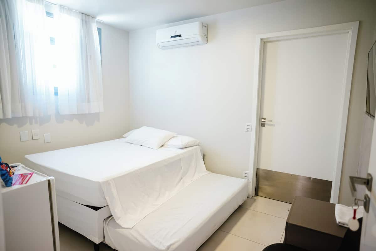 Quarto pequeno e limpo com uma cama de solteiro, uma escrivaninha, uma cadeira, uma porta e um aparelho de ar condicionado na parede.