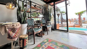 Quarto com teto de madeira, grandes janelas e vista para uma piscina. Há várias cadeiras e uma mesa sobre um tapete estampado.