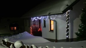 Uma casa com chão coberto de neve, cerca de madeira e luzes de cordão.
