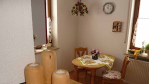 Uma área de refeições com uma mesa de madeira, cadeiras e toalha de mesa florida. Há um relógio na parede e um cesto de flores pendurado.