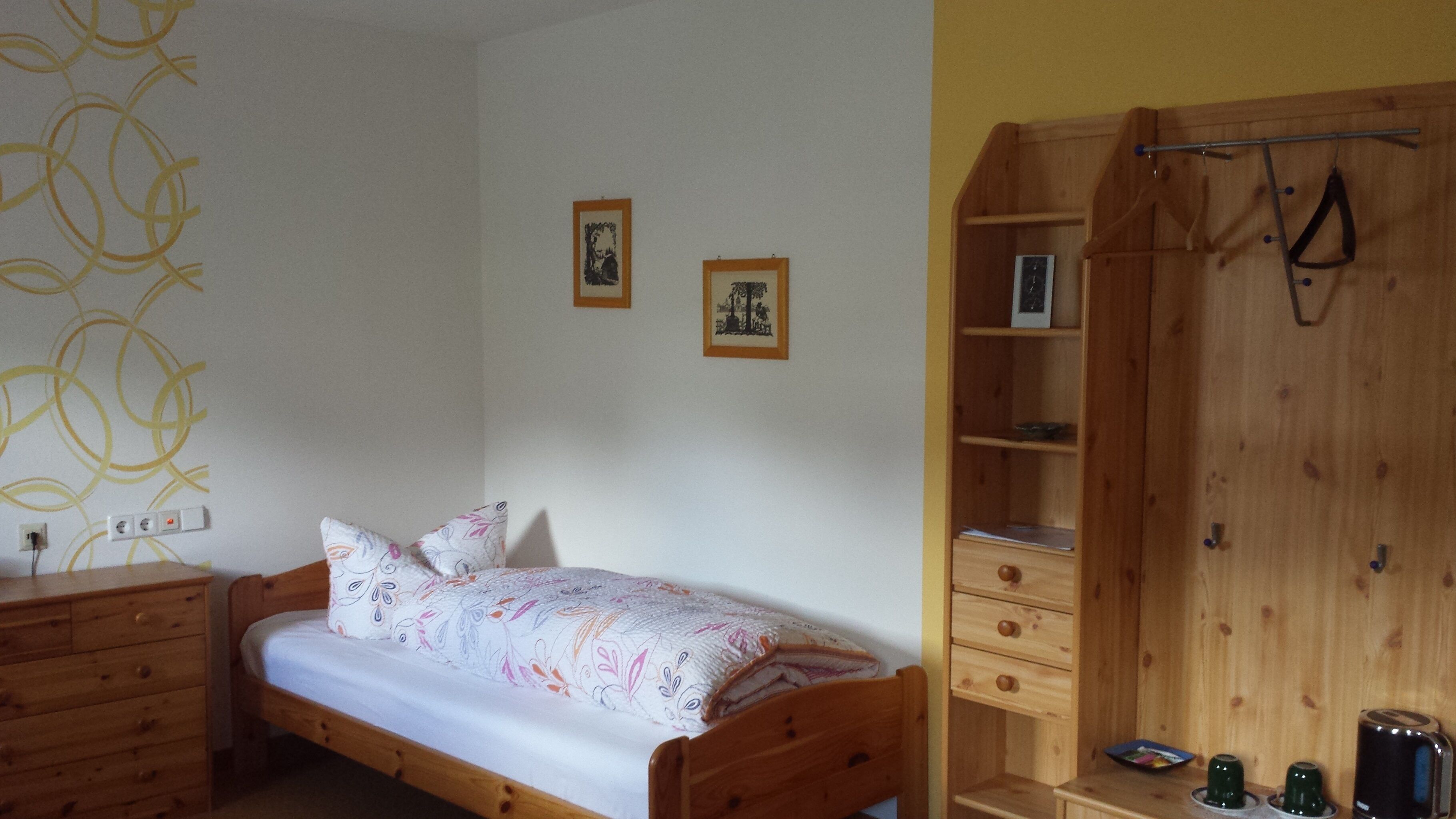Um quarto com uma cama de madeira, um guarda-roupa de madeira e uma pequena escrivaninha com cadeira. Há duas fotos emolduradas na parede.