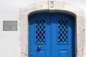 Uma porta azul com padrão decorativo na janela, um puxador de latão e uma placa acima da porta com a inscrição 'Casa Saint Jorge'.
