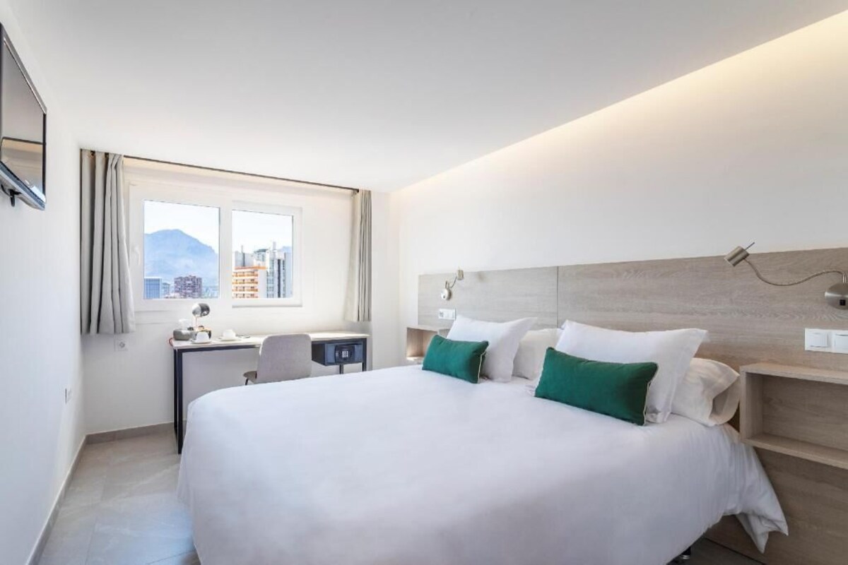 Quarto de hotel com uma cama grande, uma escrivaninha, uma cadeira, uma janela com vista para a cidade e uma televisão montada.