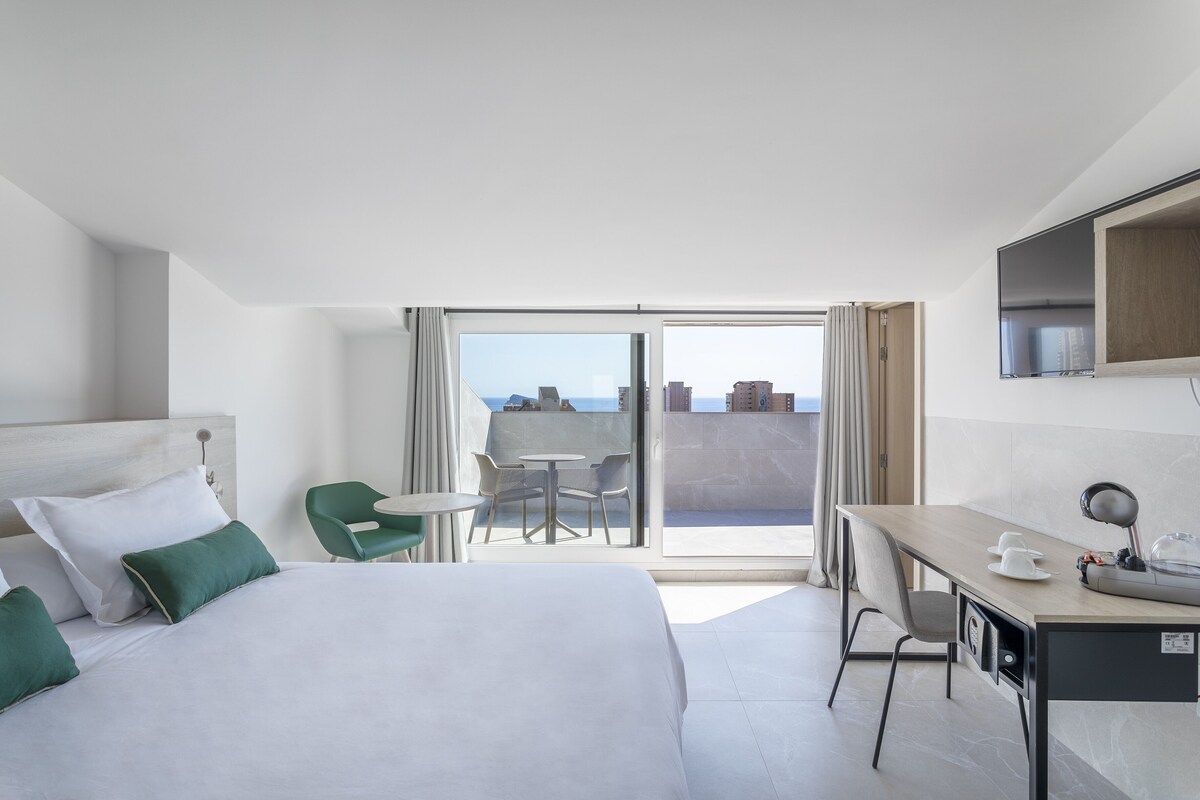 Quarto de hotel com cama, escrivaninha, cadeira, mesa e varanda com vista.