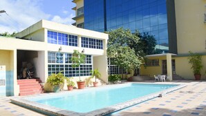 Piscina com bordas azulejadas, rodeada por vasos de plantas e um edifício com grandes janelas.