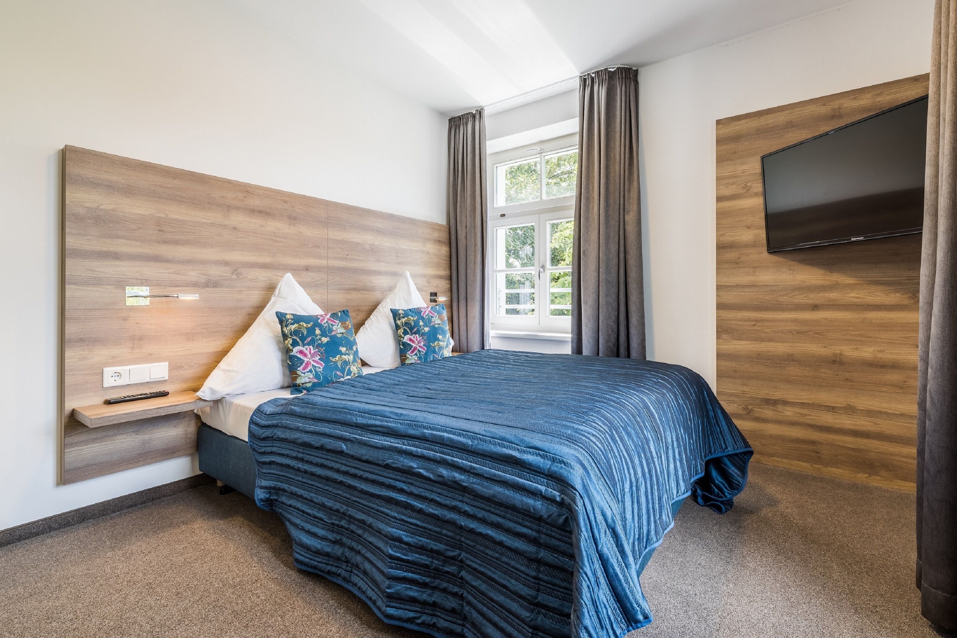 Quarto de hotel com uma cama grande, uma TV na parede, uma janela com cortinas e uma mesa de cabeceira com controle remoto.