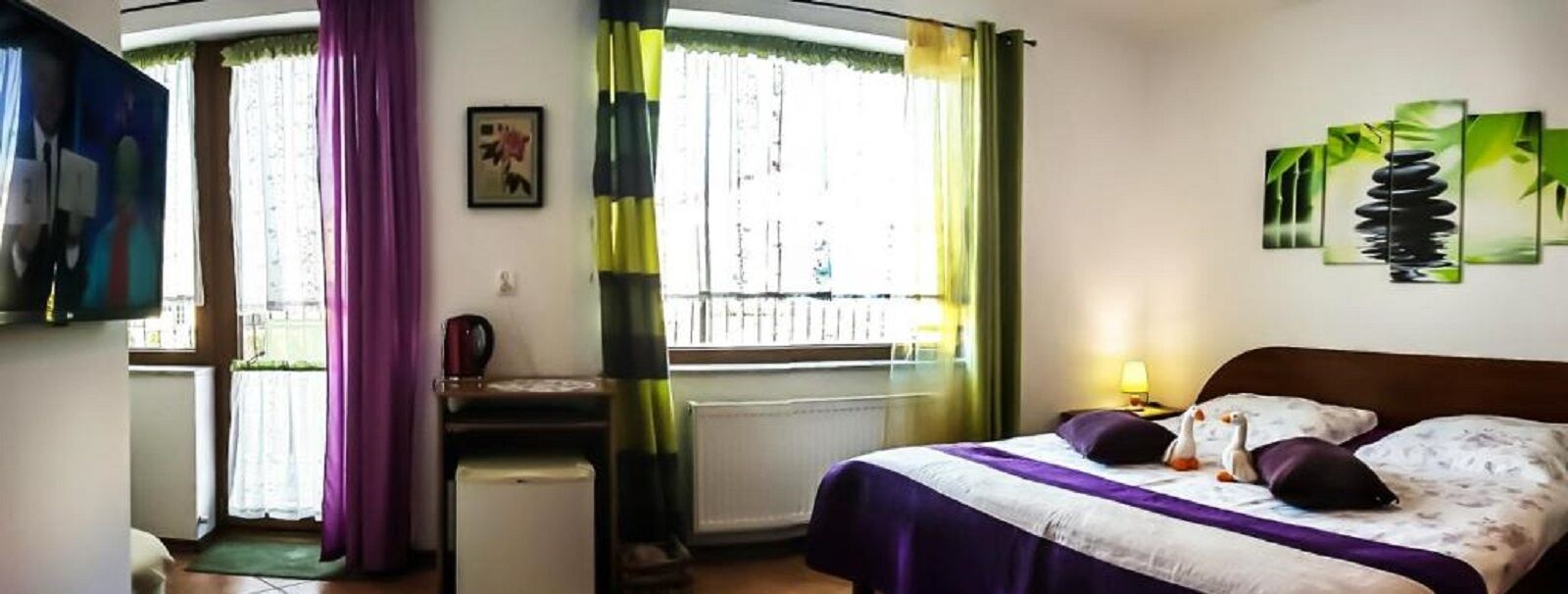Quarto com cama, radiador, janela com cortinas e televisão presa à parede.