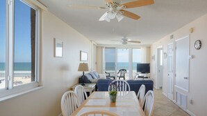 Quarto com mesa de jantar, cadeiras, ventoinha no teto, televisão e vista para a praia.