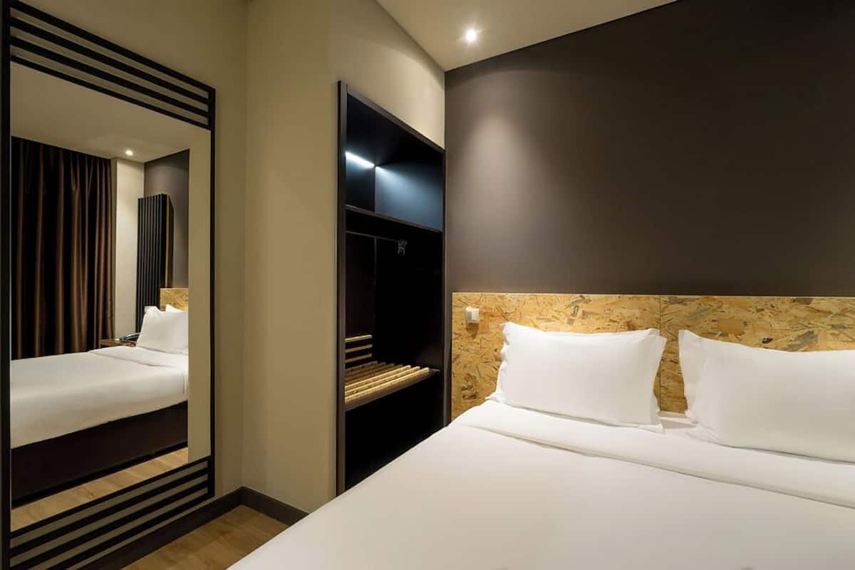 Quarto de hotel com cama, espelho, guarda-roupa e cabeceira em parede texturizada.