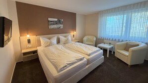 Quarto de hotel com uma cama grande, duas mesas de cabeceira, uma cadeira, uma mesinha e uma janela com cortinas finas.